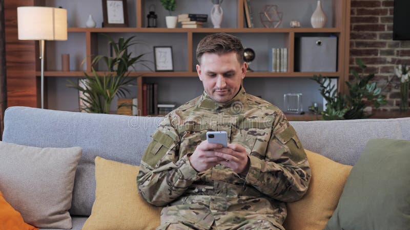 Happy Soldier in Military Uniform Sits on a Sofa at Home Using Mobile ...