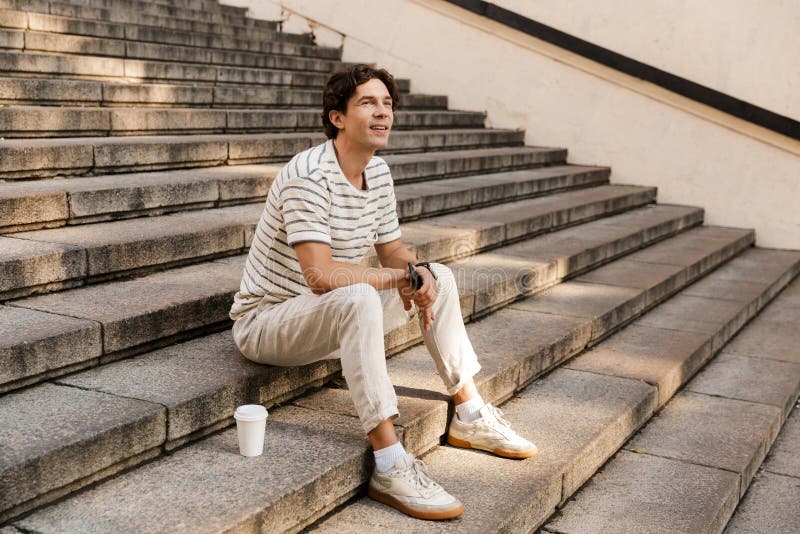 Happy Young Man Sitting on Steps Outdoors Looking Aside Holding Phone ...