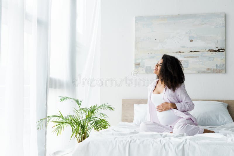 Young Pregnant African American Woman Sitting On Bed And Touching Belly