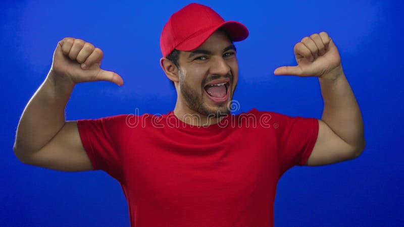 Hispanic Courier Man Wearing Red Shirt and Cap Points Thumbs in Studio with Open Mouth ...