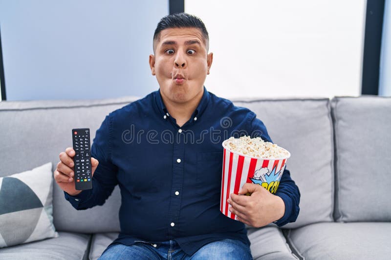 Hispanic Young Man Eating Popcorn Using Tv Control Making Fish Face with Mouth and Squinting ...
