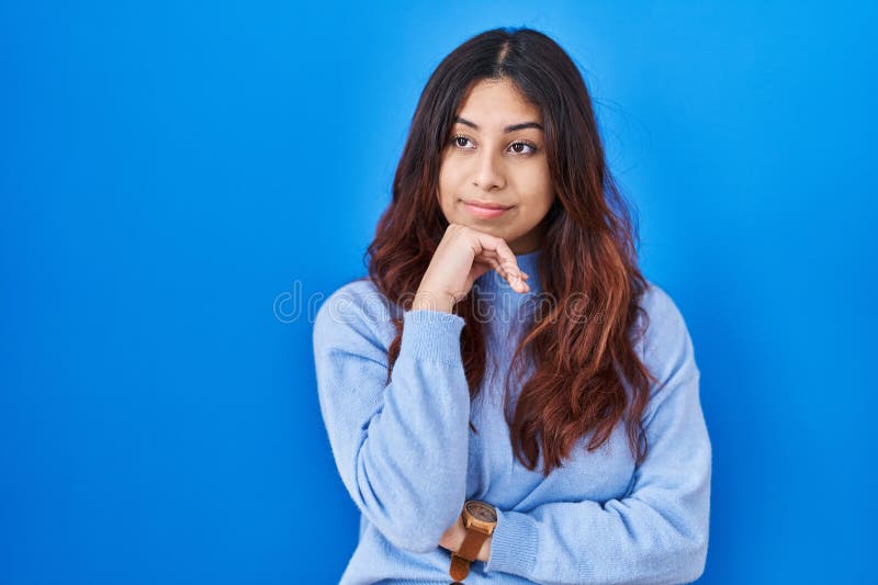 Hispanic Young Woman Standing Over Blue Background with Hand on Chin ... Hispanic Young Woman Standing Over Blue Background with Hand on Chin ...