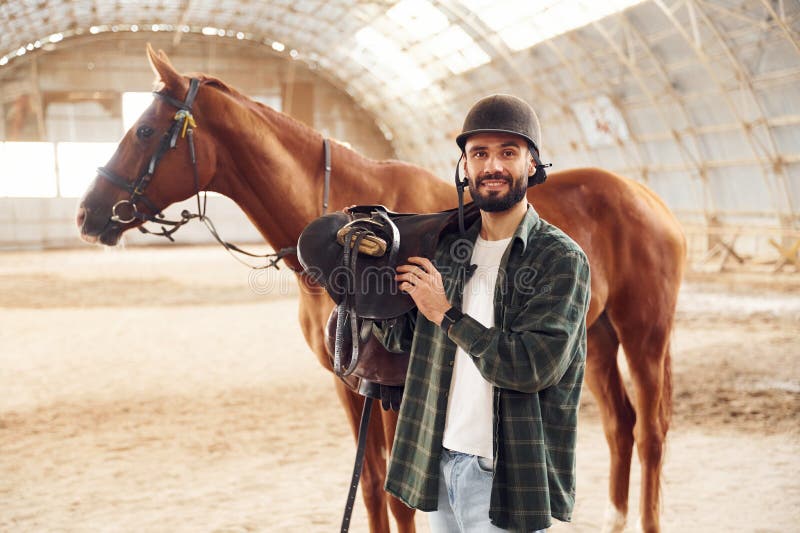 Holding the Saddle in Hands. Young Man with a Horse is in the Hangar ...