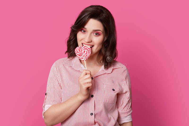 Image Of Pleased Dark Haired Female Model Bites Sweet Candy Looks At