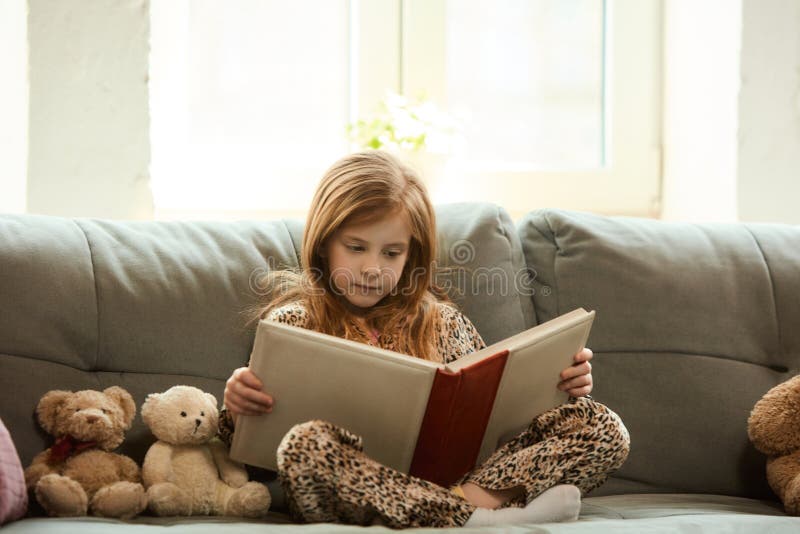 Intense Little Reader Sits on Comfortable Grey Couch, Holding Large ...