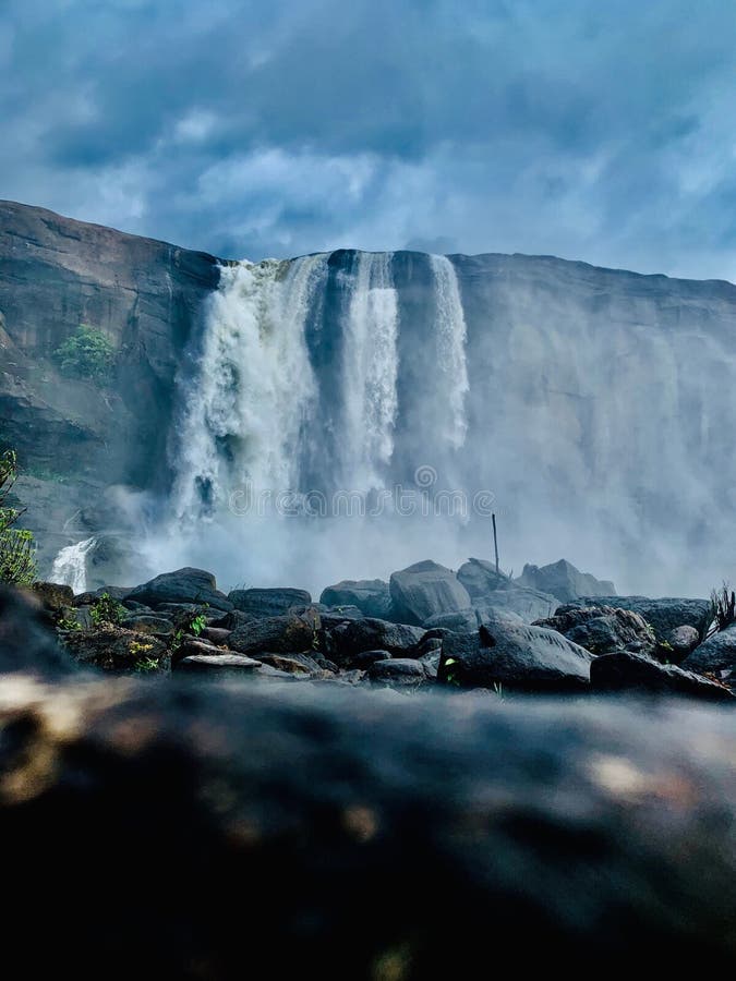 A Large Waterfall Towering Above Some Rocks Under a Cloudy Sky Stock ...