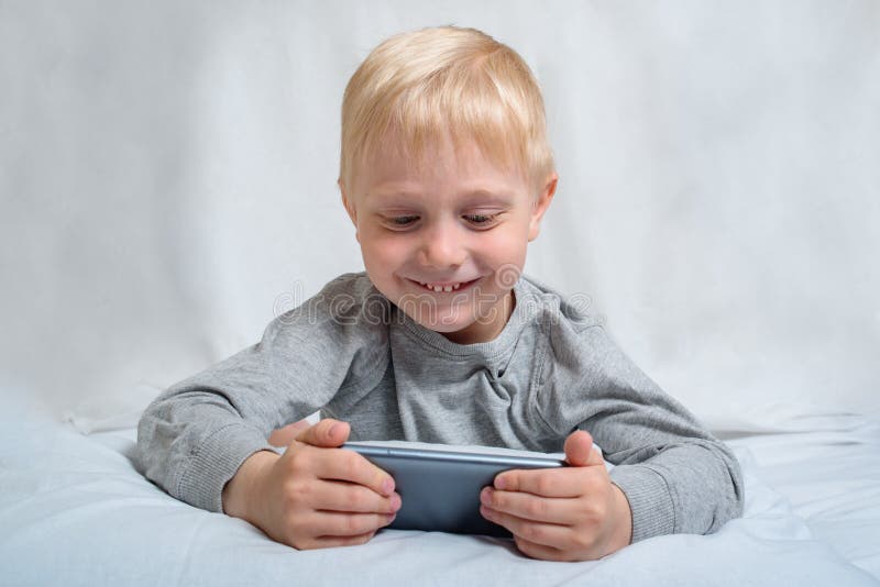 Little Blond Boy is Watching Something on a Smartphone while Lying in ...