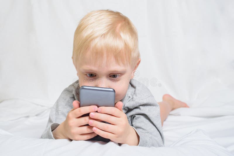 Little Blond Boy is Watching Something on a Smartphone while Lying in ...