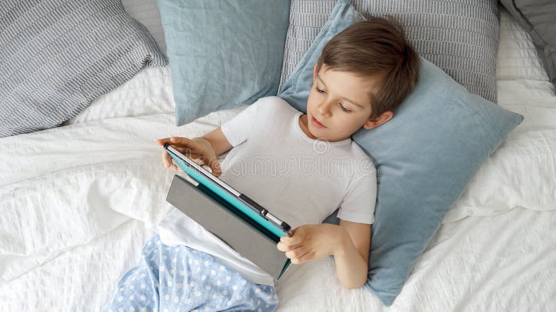 Little Boy is Lying on His Bed, Using a Tablet Computer during the Day ...