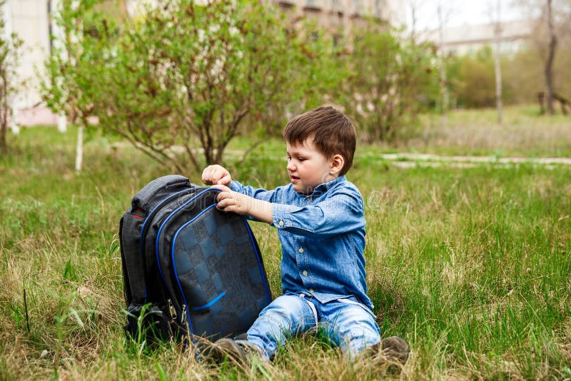 A Little Boy Opens Up His Large Backpack Sitting on the Grass Stock ...