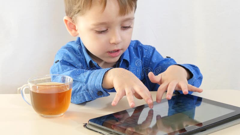 A Little Boy is Sitting at a Table and Playing on a Tablet. the Child ...