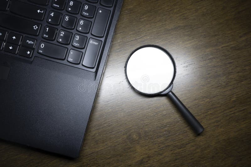 A Magnifying Glass with the Key Pad of a Laptop are on the Brown Wooden ...