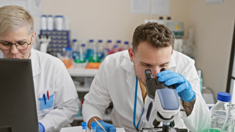 A Male Scientist Using a Microscope beside a Female Colleague, Both in ...