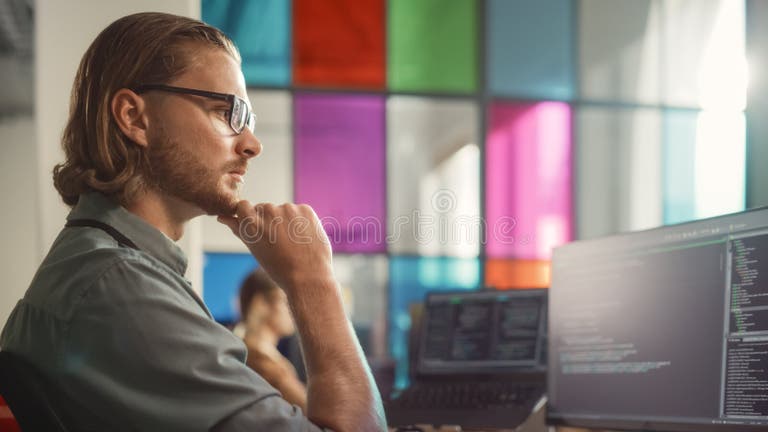 Male Senior Software Engineer Coding on Desktop PC with Multiple Monitors Setup and Laptop Aside ...