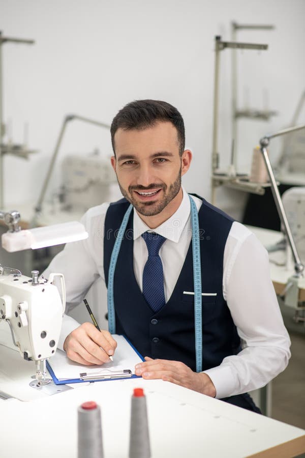 Male Tailor Sitting at Sewing Machine, Making Notes, Smiling Stock ...
