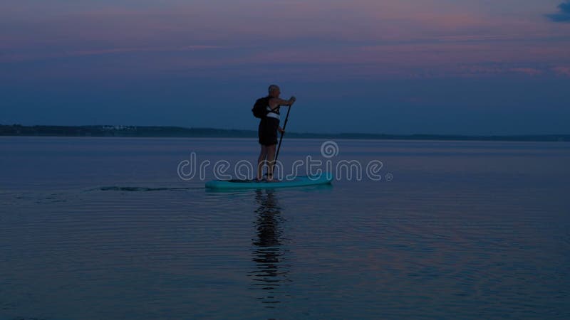 A Man with a Backpack on a SUP Board Swims in the Lake at Dusk in the ...