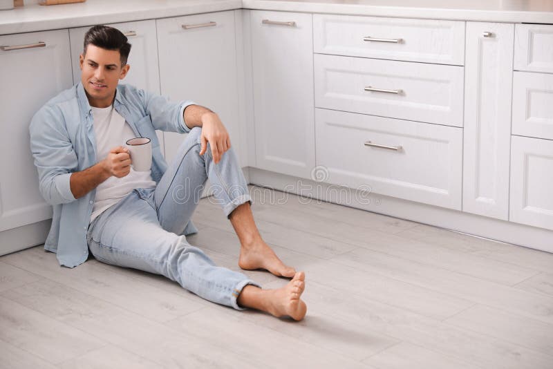 Man with Cup of Drink Sitting on Warm Floor in Kitchen, Space for Text ...