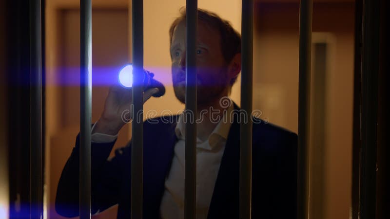 A Man with a Flashlight is Seen Behind Prison Bars at Night, Providing ...