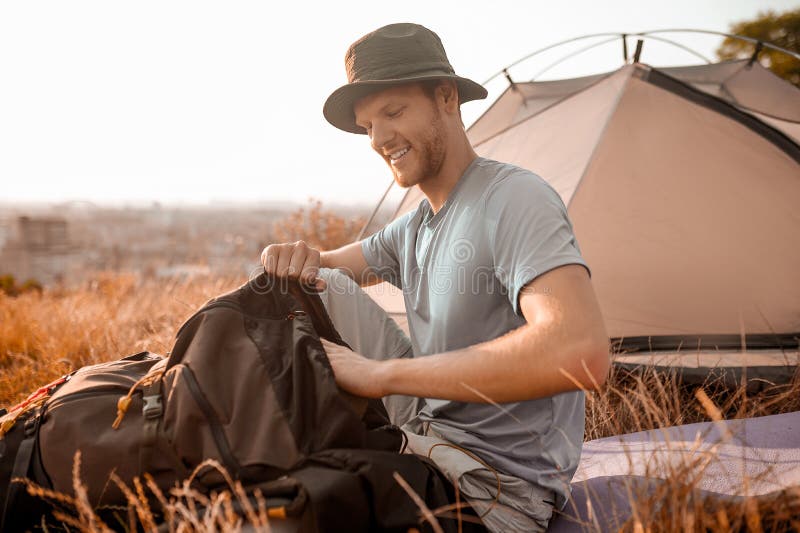 A Man in a Hat Packing His Backpack while Sitting Near the Tent Stock ...