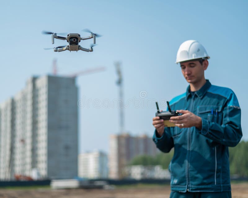 A Man in a Helmet and Overalls Controls a Drone at a Construction Site ...