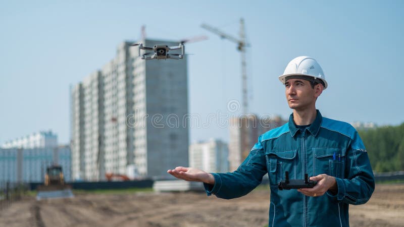 A Man in a Helmet and Overalls Controls a Drone at a Construction Site ...