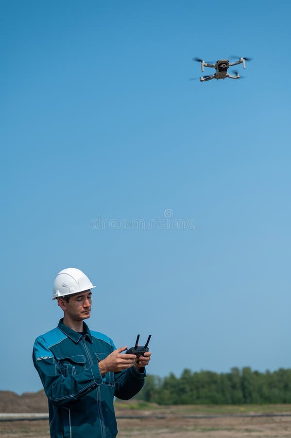 A Man in a Helmet and Overalls Controls a Drone at a Construction Site ...