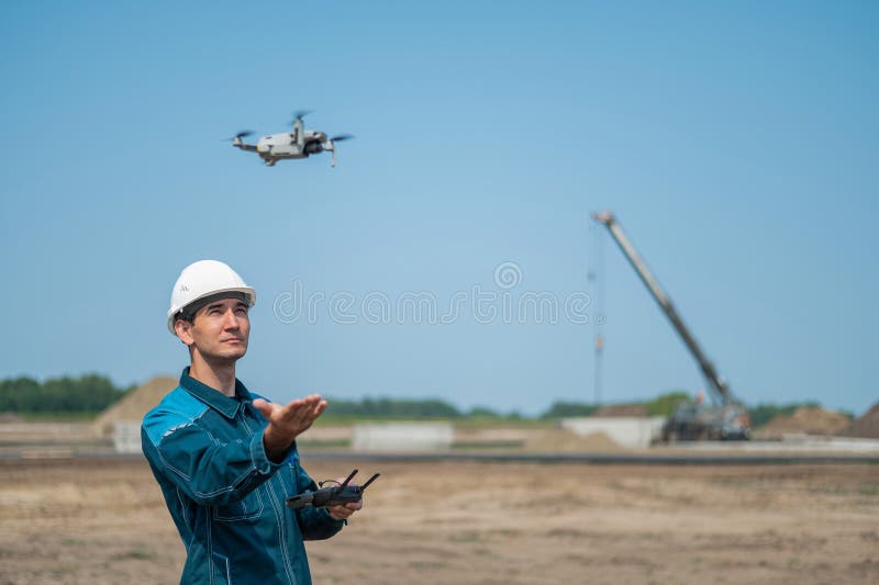 A Man in a Helmet and Overalls Controls a Drone at a Construction Site ...