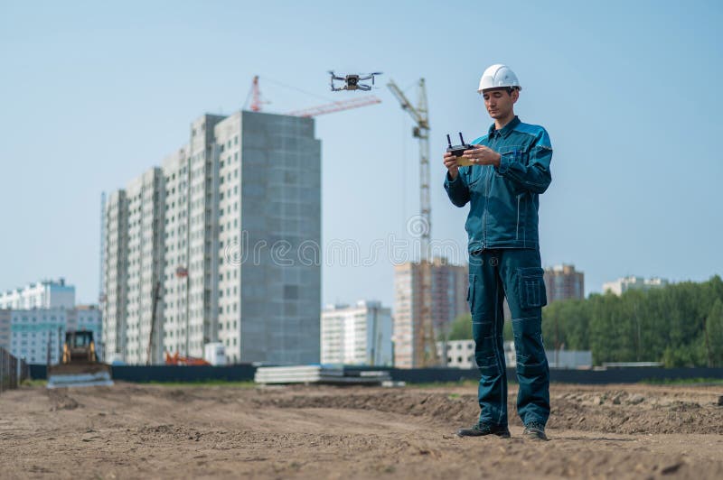 A Man in a Helmet and Overalls Controls a Drone at a Construction Site ...