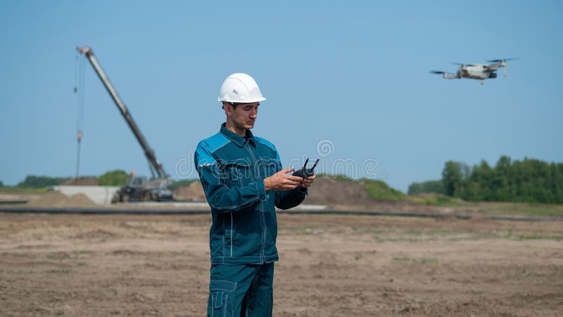 A Man in a Helmet and Overalls Controls a Drone at a Construction Site ...