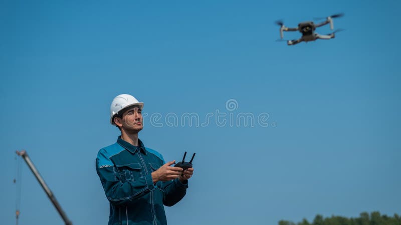 A Man in a Helmet and Overalls Controls a Drone at a Construction Site ...