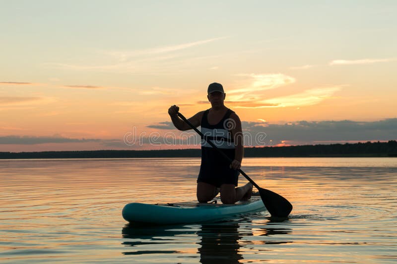 A Man In Shorts Stands On A Sup Board In The Evening At Sunset In The