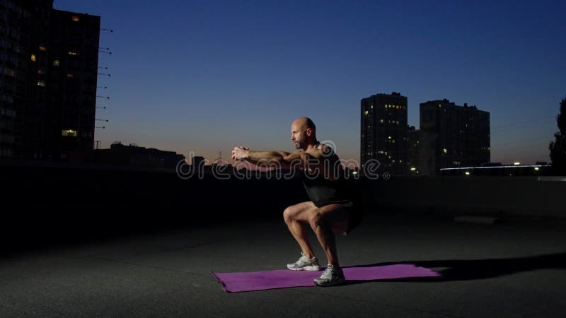 A Man Performs a Static Squat Exercise on a Roof in a Night City. Stock ...