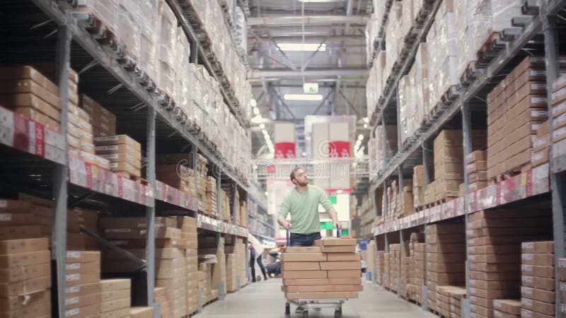 A Man is Pushing a Trolley Full of Boxes on it between Shelves with ...