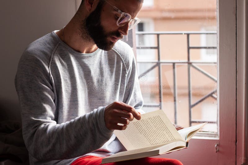 Young Man with Glasses Reading a Book Sitting Next To the Window during ...