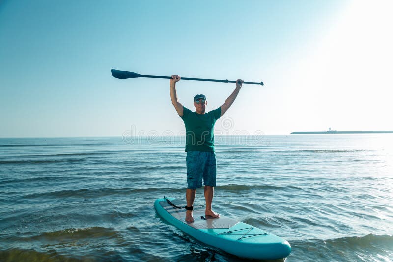 A Man In Shorts Stands On A Sup Board In The Evening At Sunset In The