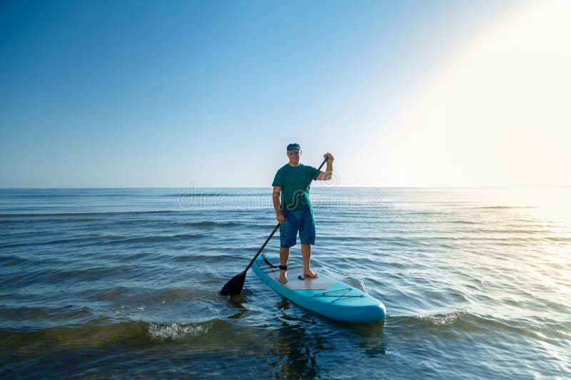 A Man In Shorts Stands On A Sup Board In The Evening At Sunset In The