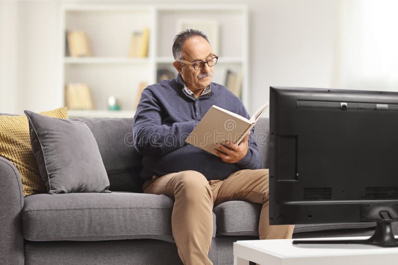 Man Sitting on a Couch and Reading a Book in Front of Tv at Home Stock ...