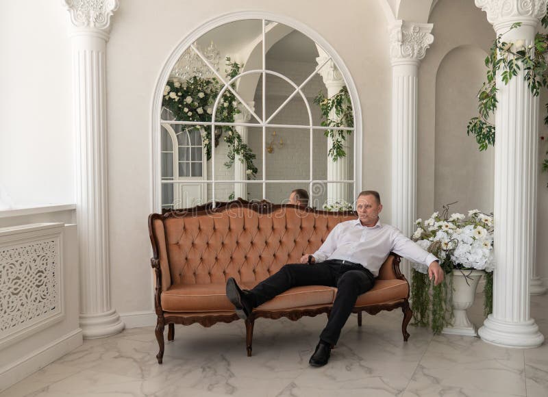 A Man is Sitting on a Sofa in a White Room with a Window Behind His ...