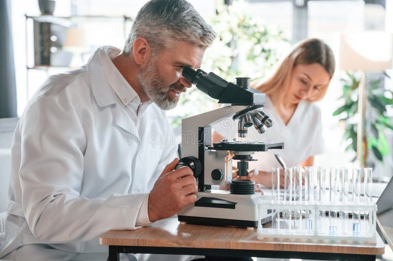 Man is Sitting and Using Microscope. Two Clinic Workers are Indoors ...