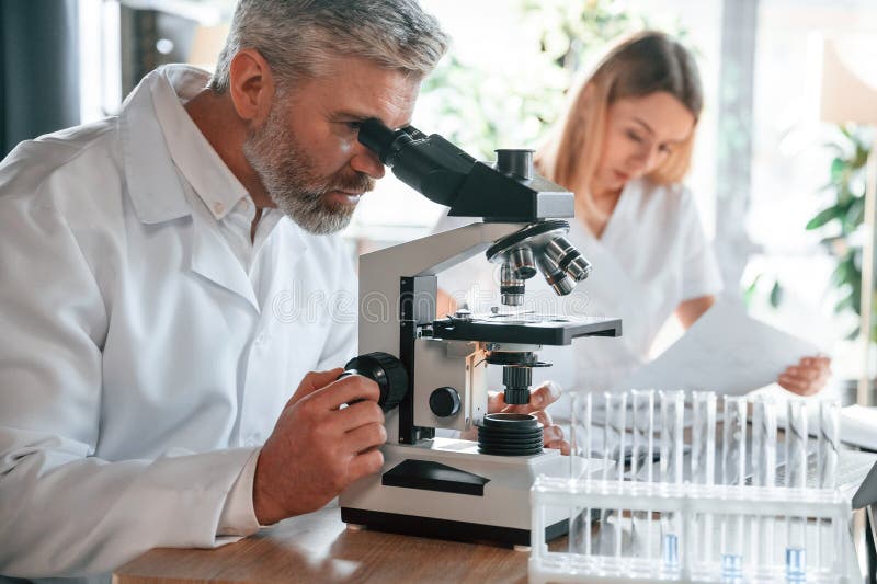 Man is Sitting and Using Microscope. Two Clinic Workers are Indoors ...