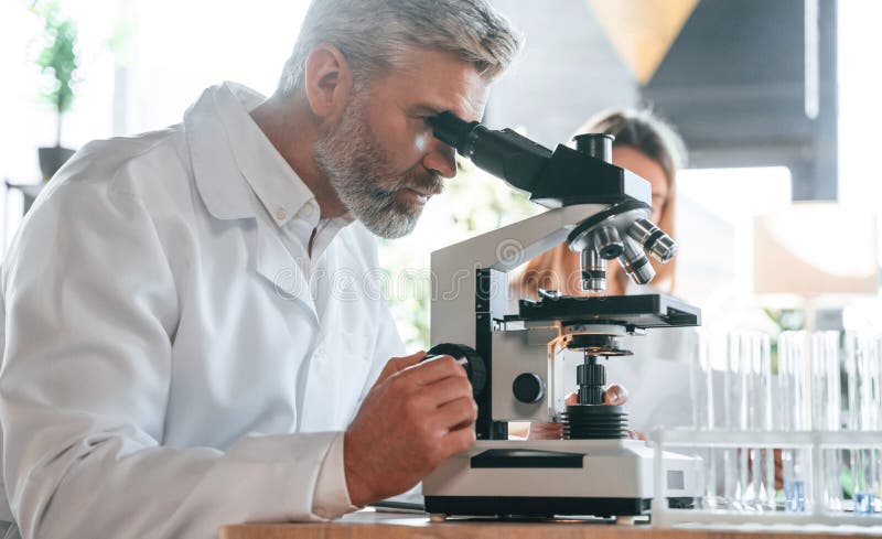 Man is Sitting and Using Microscope. Two Clinic Workers are Indoors ...