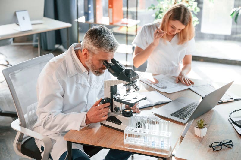 Man is Sitting and Using Microscope. Two Clinic Workers are Indoors ...
