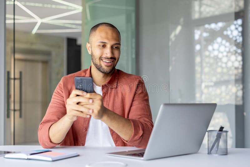 Man Smiling at Phone while Seated at Desk with Open Laptop, Showing a ...