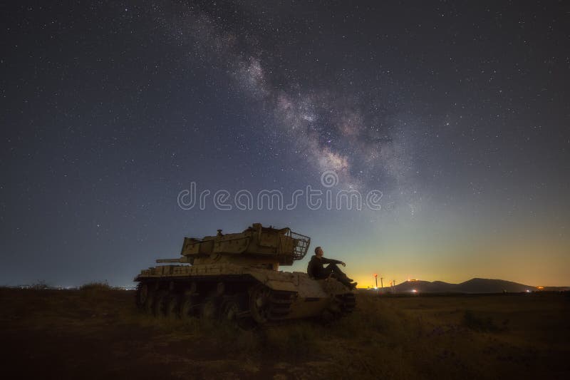 Man Stargazing while Sitting on a Rusty Tank with Mountains and Lights ...