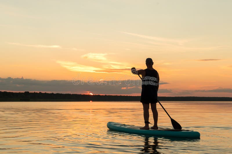 A Man In Shorts Stands On A Sup Board In The Evening At Sunset In The