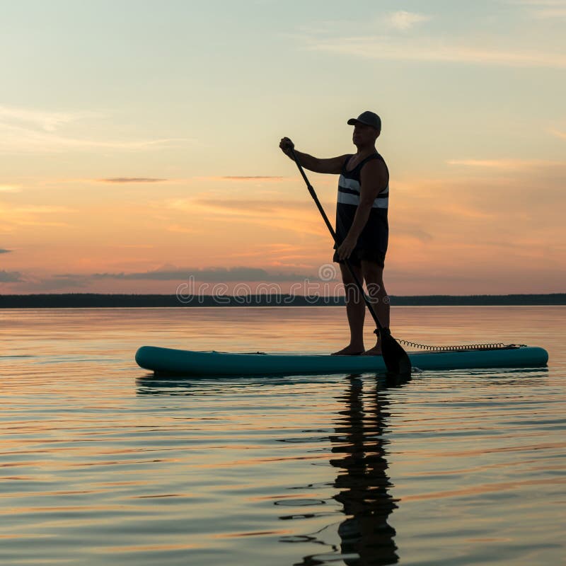 A Man In Shorts Stands On A Sup Board In The Evening At Sunset In The