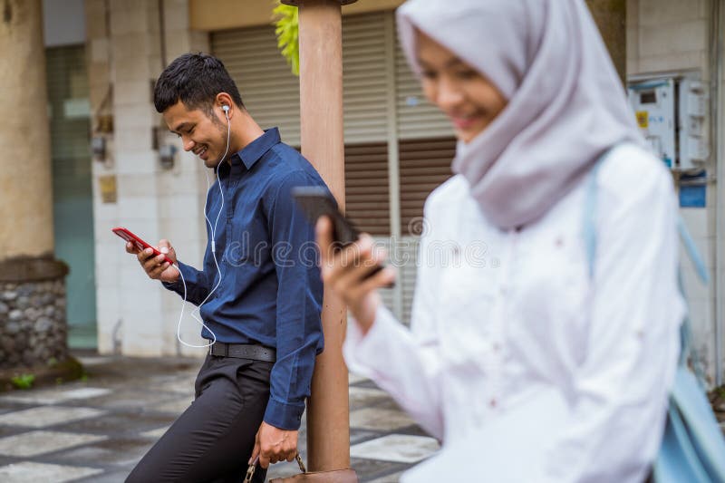 Man Wearing Earphones Using Mobile Phone and Muslimah Worker Also Stock ...