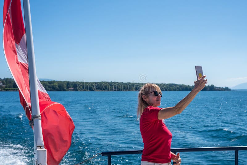 Mature Blonde Woman With Glasses Taking A Selfie On A Boat With Swiss Flag Fluttering On Lake