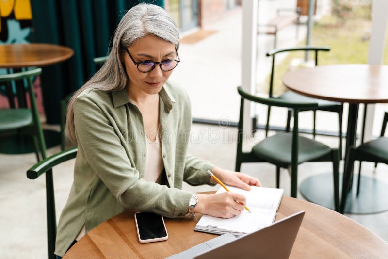 Mature Grey Woman Writing Down Notes while Working with Laptop in Cafe ...