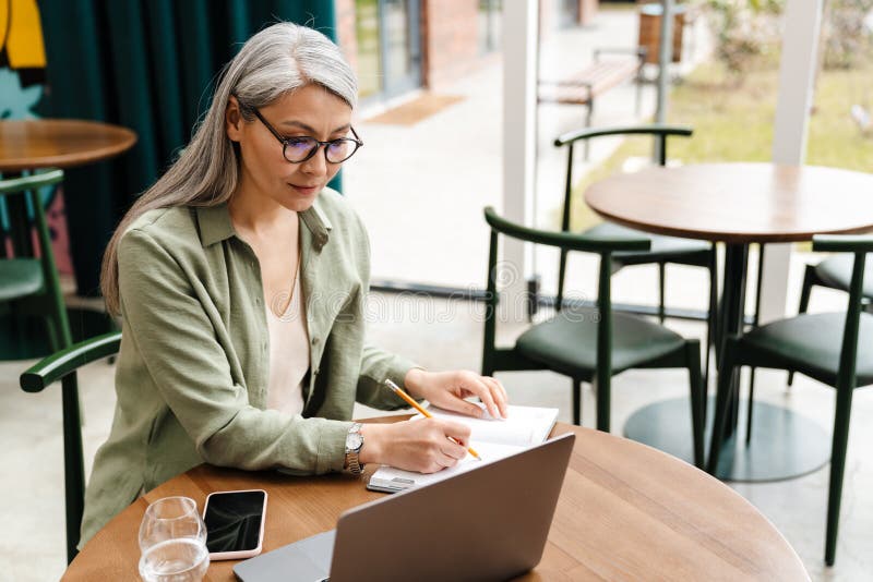 Mature Grey Woman Writing Down Notes while Working with Laptop in Cafe ...